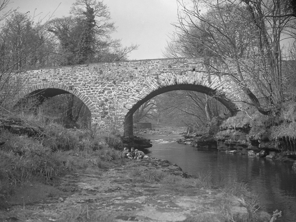 Group of boys crouching on river bank under a stone bridge